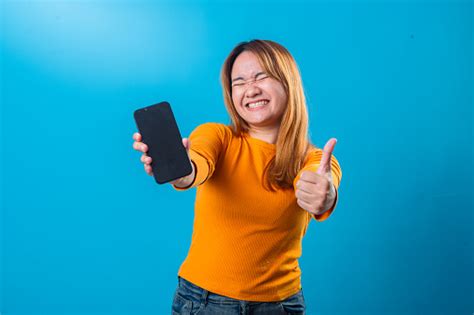 주황색 상의를 입은 아시아 여성이 스마트 폰을 들고 밝게 웃으며 단색 파란색 배경에 엄지 손가락을 치켜 올리며 스튜디오 초상화에서 흥분과 승인을 보여줍니다 20 24세에