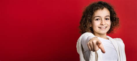 곱슬 머리 정면보기 초상화를 가진 열 살짜리 작은 백인 소녀 한 명이 빨간 배경 앞에 서서 카메라를 가리키며 행복하고 기쁨의 복사 공간을 웃고있는 손가락을 가리키고 있습니다