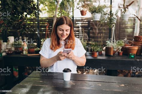 커피 종이 컵을 들고 카페에 앉아있는 동안 스마트 폰을 보는 젊은 아름다운 여성 휴대 전화를 사용하여 커피와 함께 테이블에 앉아있는 행복한 소녀 여름 시간 모바일 게임에