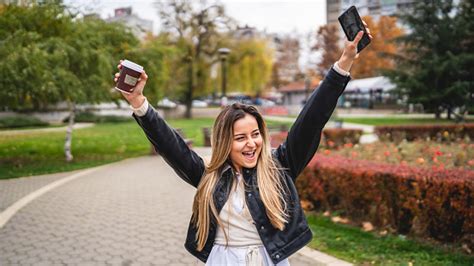 가을이나 겨울에 커피와 휴대 전화 한 잔을 들고 있는 동안 좋은 소식을 축하하는 공중에서 손을 들고 미소를 지으며 공원에 서있는 행복한 백인 젊은 여성 응원함에 대한 스톡