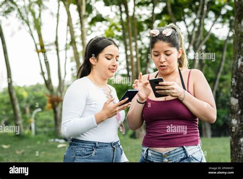 Two Latina Girls Looking At Their Cell Phones In The Park Taking A