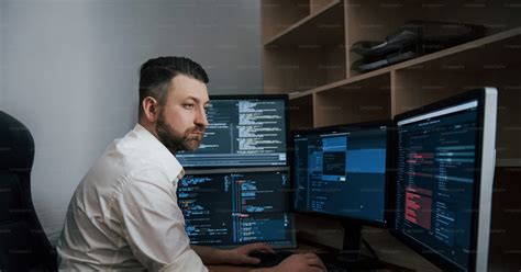 프로그램 코드는 어디에나 있습니다 흰 셔츠를 입은 수염 난 남자는 인덱스 차트에 여러 컴퓨터 화면이 있는 사무실에서 일합니다