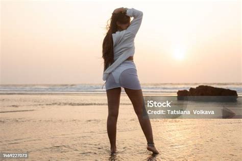 흰 수영복에 느슨한 머리를 가진 검게 그을린 날씬한 젊은 여성이 일몰을 배경으로 바닷물에 등을 대고 서 있습니다 건강한 생활