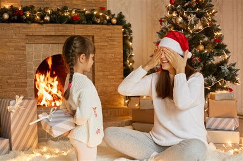 크리스마스 선물과 행복한 어머니와 아이 소녀 바닥에 앉아 손바닥으로 그녀의 눈을 덮고 깜짝 기다리고 뒤에 선물 상자와 아이 산타 클로스 모자여성 25 29세에 대한 스톡