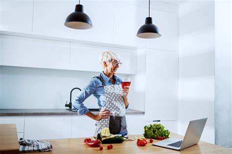 부엌에 서서 와인을 마시고 건강한 저녁 식사를 준비하는 앞치마에 미소 백인 선임 여성 주방 카운터에는 고추 양상추 체리 토마토 및 노트북이 있습니다 가정 생활에 대한 스톡