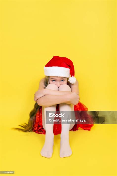 빨간 드레스와 산타 클로스 빨간 모자를 쓴 슬픈 십대 소녀가 앉아서 무릎을 껴안습니다 노란색 배경에 고립 된 산타 모자의 소녀