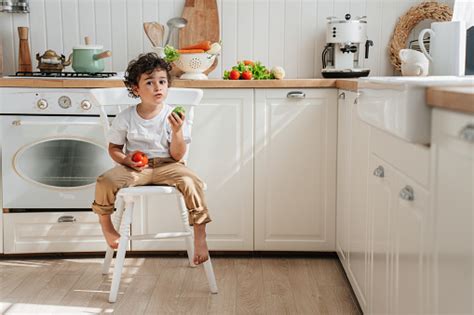 토마토를 들고 오이를 먹는 바지를 입은 귀여운 백인 소년 건강 한 음식 개념입니다 부엌에서 곱슬 스페인 잘 생긴 아이는 야채를
