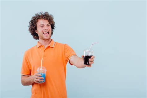 곱슬 머리 미소와 잘 생긴 행복 소년 오렌지 셔츠 고립 된 파란색 배경 긍정적 인 얼굴 감정 복사 공간을 입고 주스와 두 개의 플라스틱 컵을 보유하고 사람들에 대한 스톡