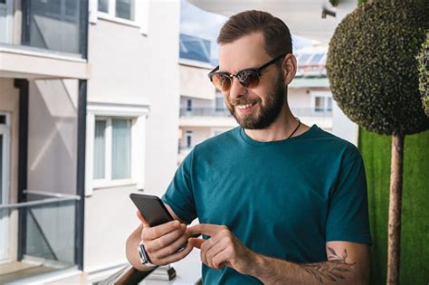 캐주얼하게 옷을 입은 남성 관광객이 스마트 폰을 탐색하고 호텔 방의 테라스에 서있는 동안 온라인으로 뉴스를 확인하는 젊은 30 39세에 대한 스톡 사진 및 기타 이미지