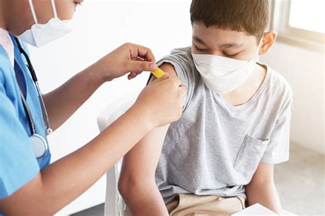 아프리카 의사 또는 간호사 여성이 예방 접종을하고 코카서스 소년이 병원에서 마스크를 착용하고 있는 석고를 덮고 있습니다