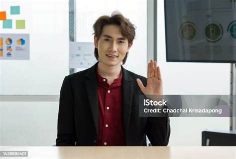 검은 양복을 입은 젊은 아시아 사업가가 책상에 앉아 휴식을 취하고 앞에서 손을 흔든다 배경에는 유리판에 메모지와 문서가 있었다 25 29세에 대한 스톡 사진 및 기타 이미지