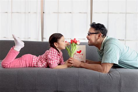 행복한 아버지의 날 교정을 위해 거실의 소파에서 어린 아빠에게 수제 인사말 카드와 꽃을주는 사랑스러운 귀여운 소녀 행복한 가족