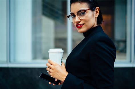 도심에서 커피 휴식 중 카메라를 보고 성공적인 여성 전문가의 절반 길이 초상화 도시 설정에 서 손에 새로운 휴대 전화와 긍정적 인 지능형 여성 상인 거리에 대한 스톡 사진