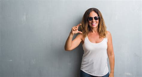 중세 스페인 여자 선글라스 미소와 자신감이 생기고 카메라 동안 손가락으로 크기 표시를 하 고 손으로 몸짓을 입고 회색 벽에 측정 개념입니다 갈색 머리에 대한 스톡 사진 및