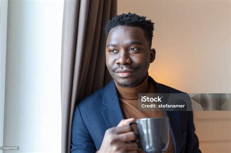 호텔 방에서 커피를 마시는 창가에 서있는 사업가 사려 깊은 젊은 아프리카 사업가커피 한 잔을 들고 창가에 서서 밖을 바라보고 있습니다 20 24세에 대한 스톡 사진 및 기타