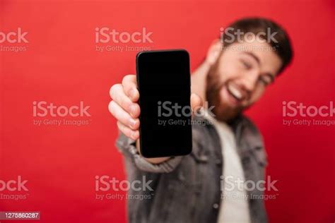 카메라와 미소 빨간 배경에 고립 된 미소의 초점을 맞춘 검은 화면을 보여주는 청바지 재킷에 수염을 가진 즐거운 남자의 사진 가리키기에 대한 스톡 사진 및 기타 이미지