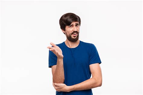 파란 셔츠를 입은 잘생긴 수염을 가진 브루넷 남자가 팔을 가슴에 접고 한 손을 공중에 던지며 하얀 배경 위에 고립된 회의적인