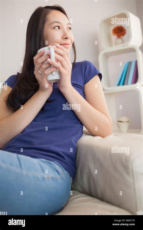 Thoughtful Young Asian Woman Sitting On The Couch Holding Mug Stock