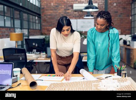 Biracial And African American Young Female Architects Discussing Over Blueprint At Desk In
