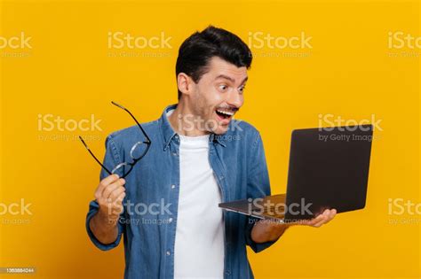 깜짝 놀랄 행복 행복 한 행복 한 흥분 자신감 면도 백인 남자 안경을 벗고 그의 손에 오픈 노트북을 들고 고립 된 오렌지 배경에 서 있는 동안 화면에 놀라움과 행복 하 게