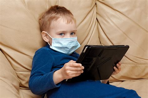 의료 용 마스크를 쓴 귀여운 아기 소년은 태블릿 패드를 손에 들고 가죽 소파에 앉아 있습니다 파란 잠옷을 입고 감성적인 얼굴