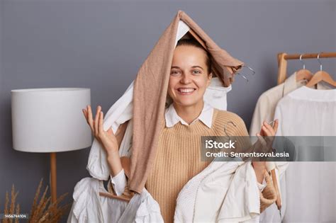옷 근처에 서 있는 쾌활한 웃긴 여자의 실내 사진은 머리에 베이지색 점퍼를 입고 선반에 걸려 있고 옷걸이에 옷을 걸고 패션 매장에서 쇼핑을 즐기고 있습니다 30 34세에