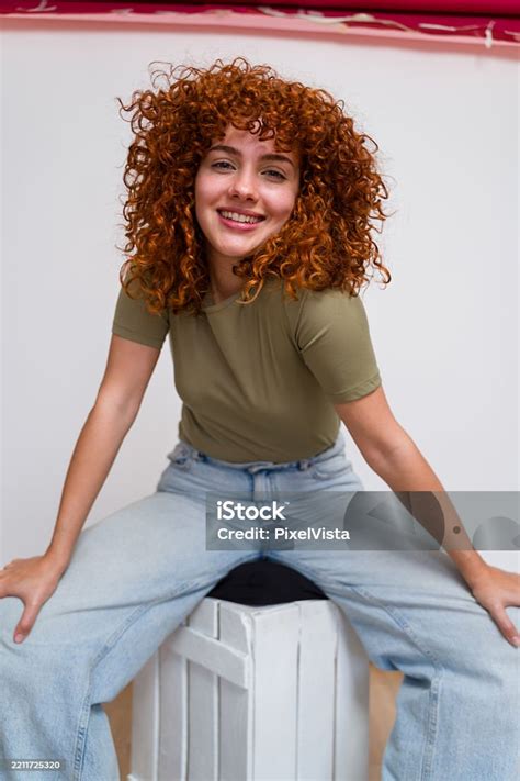빨간 곱슬 머리를 한 젊은 여자가 하얀 나무 상자에 미소 짓고 앉아 있습니다 20 29세에 대한 스톡 사진 및 기타 이미지 20 29세 T 셔츠 곱슬 머리 Istock