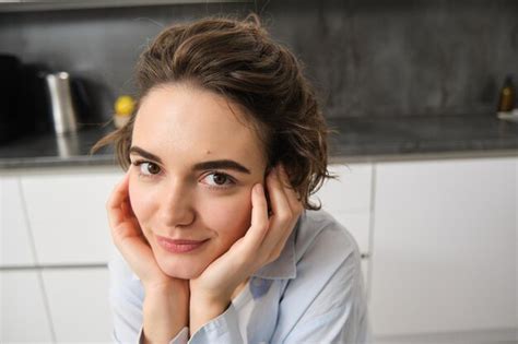 Feche O Retrato De Uma Jovem Morena Que Olha Para A C Mera E Sorri Sentada Em Casa Na Cozinha