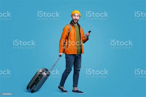 캐주얼하게 옷을 입은 젊은 수염을 기린 남자 관광객의 전체 길이 초상화가 배낭 기내 수하물 가방 및 휴대 전화를 손에 들고 공항 터미널에서 걷고 카메라를 직접 바라보고