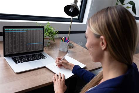 Caucasian Female Programmer Sitting At Desk Using Laptop With Coding On Screen Making Notes