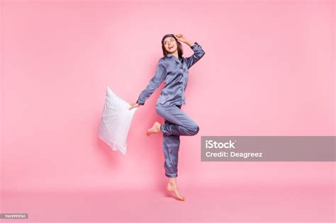 파스텔 핑크 컬러 배경에 고립 된 빈 공간을 찾고있는 베개 점프를 유지하는 잠옷을 입은 소녀의 전체 길이 신체 크기 사진