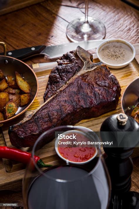 향신료 마늘 후추 로즈마리 빨간색과 흰색 소스 칼 붙이가 달린 나무 보드에 튀긴 감자 적포도주 잔 후추 통 소금 통을 곁들인 뼈에 구운 쇠고기 스테이크 수직 방향 0명에