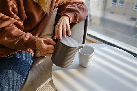 소녀는 찻주전자에서 컵에 뜨거운 차를 붓습니다 가정의 안락함을 즐기십시오 창문에서 태양광선에 있는 한 여성의 손에 하얀 컵을 클로즈업합니다 여자의 손에 차 한 잔 가정