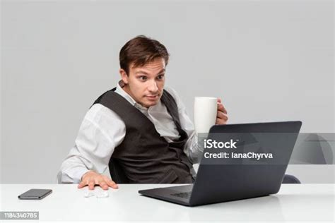 흰색 티셔츠와 회색 조끼를 입은 사무직 근로자가 커피를 마시는 흰색 테이블에 앉아 있는 노트북에 대한 정보를 확인하고 충격을 받습니다 T 셔츠에 대한 스톡 사진 및 기타
