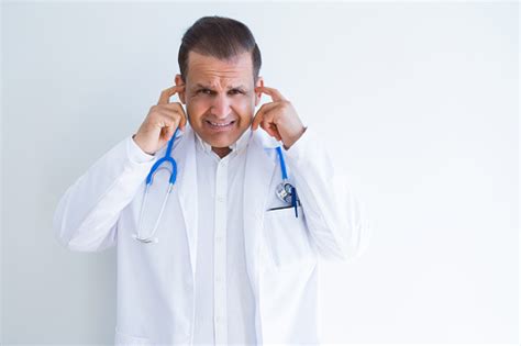 큰 소리로 음악의 소음에 대 한 짜증 표현과 손가락으로 귀를 덮고 흰색 배경 위에 청진 기 및 의료 코트를 착용 하는 중간 나이 의사 남자 청각 장애 개념 가리기에 대한