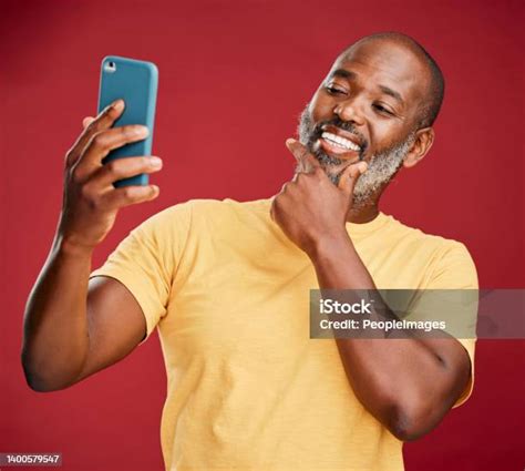 한 관상적인 트렌디 한 성숙한 아프리카 계 미국인 남자가 빨간 스튜디오 배경에 대해 스마트 폰에서 셀카를 찍습니다 소셜 미디어에 사진을 찍는 동안 서서 포즈를 취하는 세련된