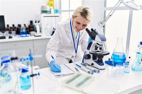 Young Blonde Woman Scientist Using Microscope Taking Notes At
