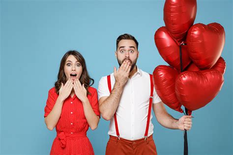 생일 휴가 파티를 축하하는 흰색 빨간 옷을 입은 충격적인 젊은 부부 친구 남자 여성은 파란색 배경에 고립 된 손으로 입을 덮는 공기 팽창 헬륨 풍선무리를 보유하고 있습니다