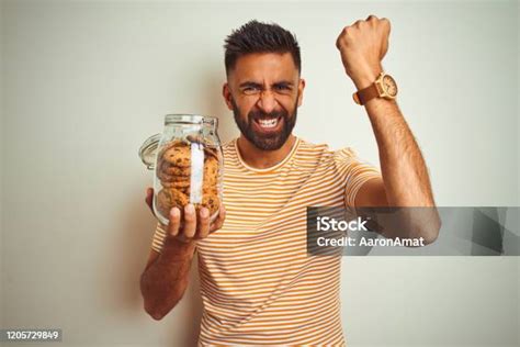 고립 된 흰색 배경 위에 서있는 쿠키 항아리를 들고있는 젊은 인도 남자는 화가 나고 화가 나서 분노로 외치고 미친 듯이 소리지르고 분노 개념으로 감정에 대한 스톡 사진 및