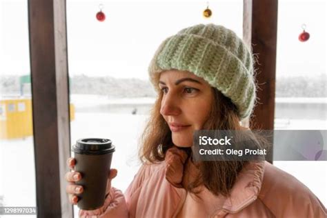 검은 종이 커피 컵을 들고 부드럽게 웃고 카페에 혼자 서서 손에 커피를 마시고 기분이 좋은 기분을 가진 것을 기쁘게 생각합니다 창 뒤의 겨울 갈색 머리에 대한 스톡 사진 및