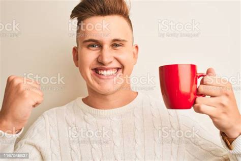 고립된 흰색 배경 위에 서있는 커피의 빨간 컵을 마시는 젊은 잘생긴 남자는 자랑스럽게 비명을 지르고 승리와 성공을 축하하는 매우 흥분 응원 감정 감정에 대한 스톡 사진 및