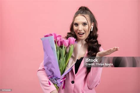 곱슬 머리로 꽃을 킁킁 거리며 즐거운 향기를 즐기고 방에서 세련된 옷을 입고 서있는 여성의 스튜디오 샷 소녀는 튤립 놀란 얼굴의 꽃다발을 들고 있습니다 20 24세에 대한