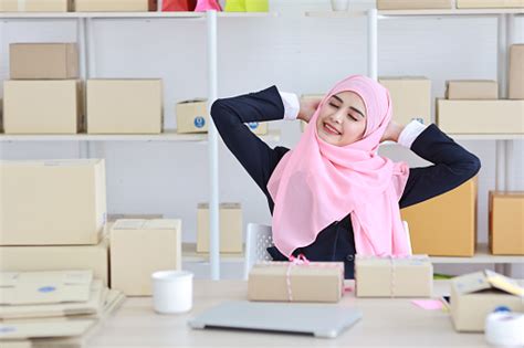 파란색 무슬림 정장을 입은 활동적인 아시아 여성이 컴퓨터와 온라인 패키지 상자 배달 작업을 한 후 앉아서 스트레칭을 하고 있다 스타트업 중소기업 프리랜서 일하는 소녀 개념