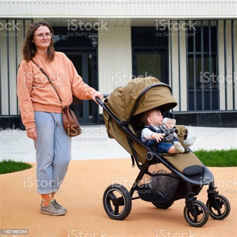 여자 어머니는 공원의 놀이터에있는 유모차에 앉아있는 유아 아기 소년과 함께 걷습니다 엄마와 함께 유모차에있는 아이 일년 된 아이 유모차에 대한 스톡 사진 및 기타 이미지