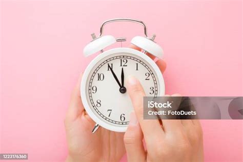 흰색 알람 시계를 들고 있는 청녀의 손 12시 방향의 화살표를 가리키는 손가락 밝은 분홍색 테이블 배경입니다 파스텔 색상입니다 시간 변경 개념입니다 근접 촬영 하향하 보기