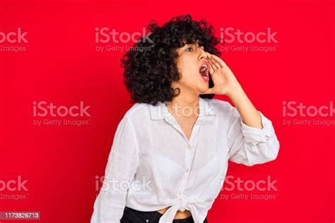 곱슬 머리를 가진 젊은 아랍 여성은 고립 된 빨간 배경 에 흰색 캐주얼 셔츠를 입고 외치고 입에 손으로 옆으로 큰 소리로 비명을 지른다 통신 개념입니다 감정에 대한 스톡