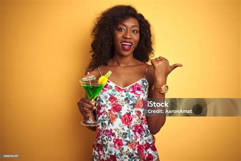 젊은 아프리카 계 미국인 여자 마시는 칵테일 위에 서 고립 된 노란색 배경 가리키는 하 고 행복 한 얼굴 미소와 함께 측면에 엄지 손가락으로 표시 가리키기에 대한 스톡 사진