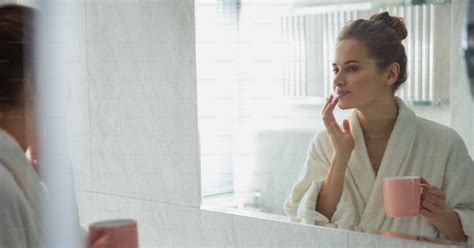 자기 관리 개념 하얀 목욕 가운을 입은 젊은 웃는 여자가 커피 한 잔과 함께 아침을 즐기고 거울 앞에서 부드럽게 입술을 만지는