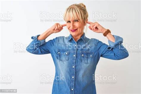 시끄러운 음악의 소음에 대한 성가신 표정으로 손가락으로 귀를 덮는 고립 된 흰색 배경 위에 서있는 캐주얼 한 데님 셔츠를 입은