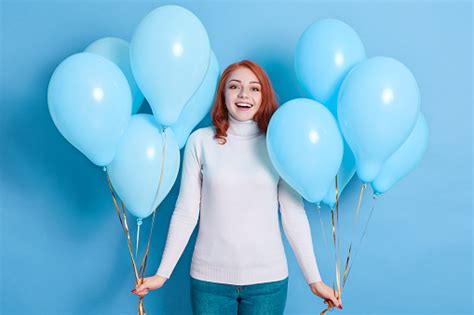 스웨터와 청바지를 입은 어린 소녀는 파란 배경에 고립 된 포즈를 취하고 널리 열린 입과 흥분된 표정으로 풍선 두 무리를 들고 행복해 보입니다 20 24세에 대한 스톡 사진